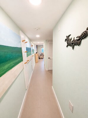 Um corredor com um quadro de paisagem na parede esquerda, uma peça decorativa de metal na direita e uma porta no final.