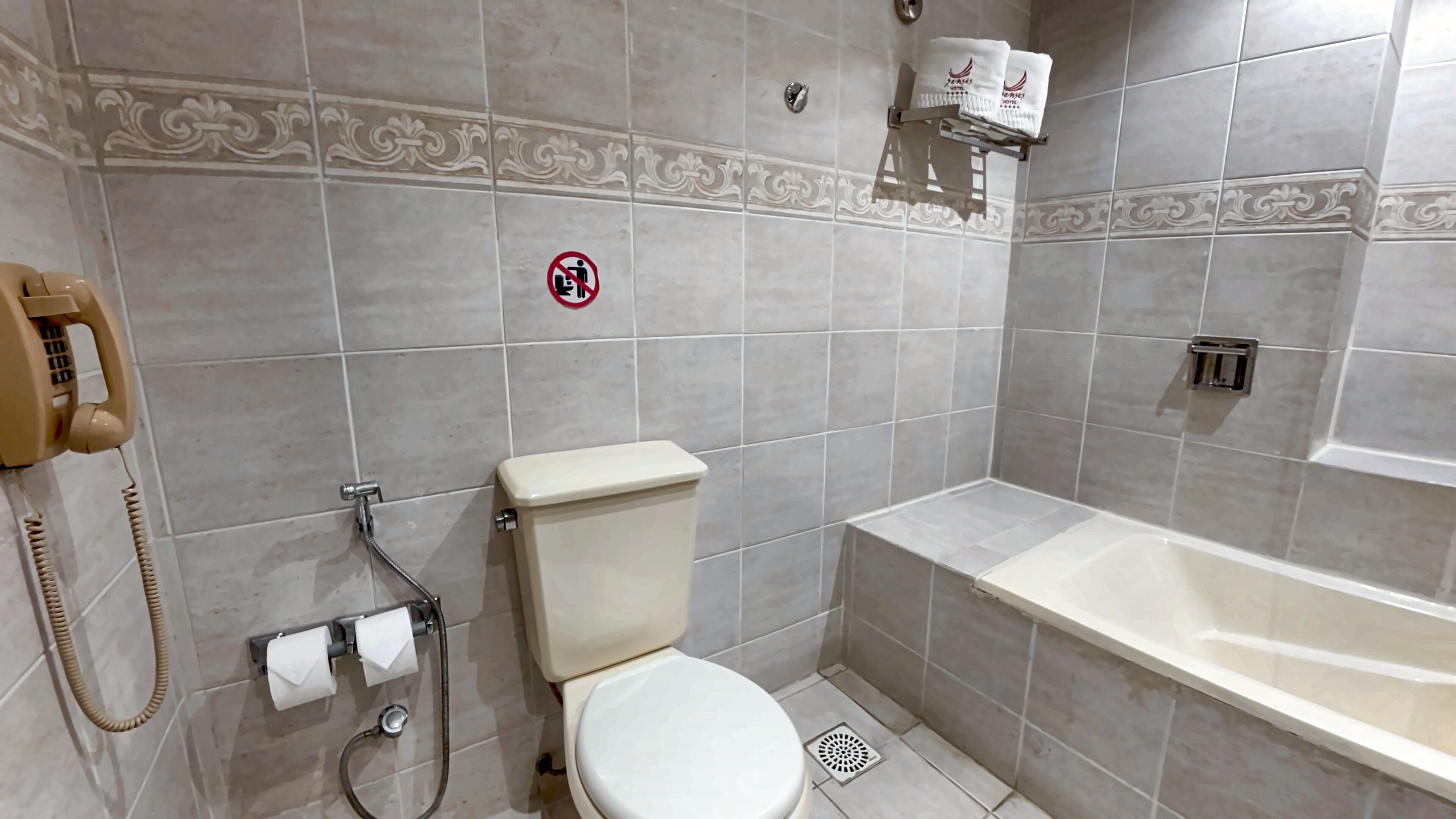 Casa de banho com sanita, bidé e banheira. Há um porta-toalhas com toalhas e um aviso de proibição de fumar na parede.