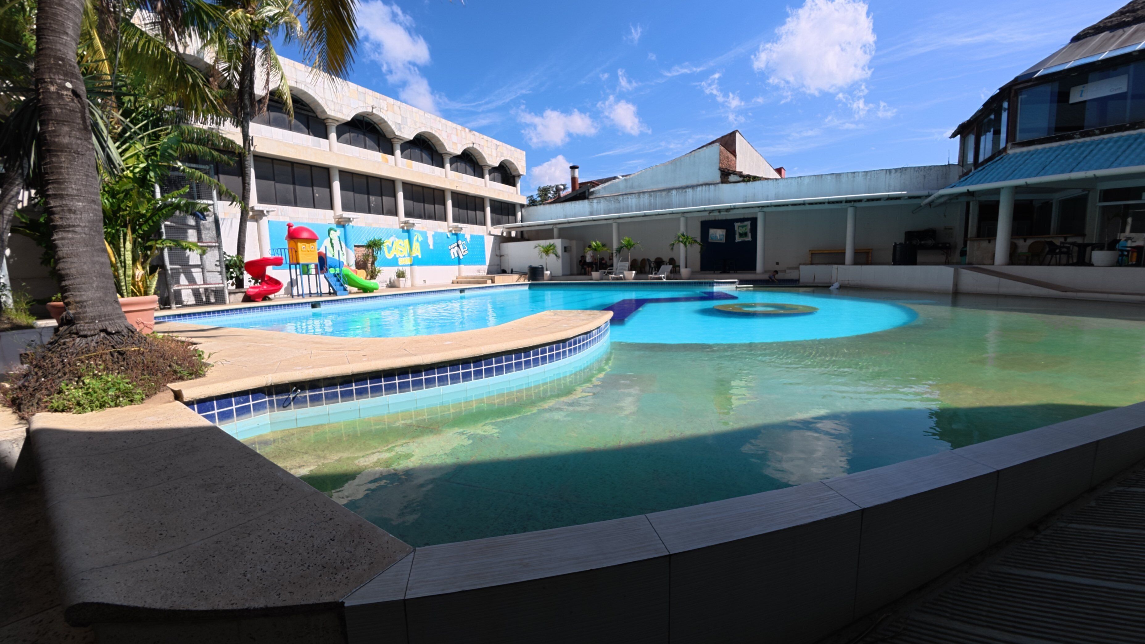 Piscina com área de recreação infantil, edifício com um mural e palmeiras.