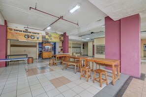 Uma cafetaria com várias mesas e cadeiras de madeira, uma máquina de vendas automática e uma entrada com persianas fechadas.