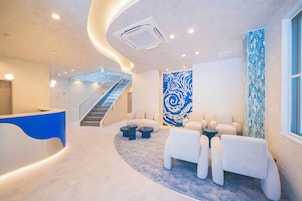 Um lobby moderno com um balcão de recepção curvo, uma escadaria, uma parede azul abstrata e uma área de estar com sofás brancos e mesas redondas.