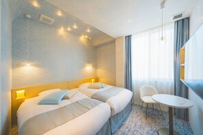 Quarto de hotel com duas camas, uma mesinha, uma cadeira e janela com cortinas.