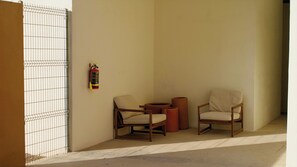 Canto de um quarto com duas cadeiras, uma mesa pequena e um extintor de incêndio na parede.