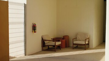 Canto de um quarto com duas cadeiras, uma mesa pequena e um extintor de incêndio na parede.
