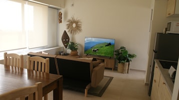 Uma sala de estar com uma mesa de jantar de madeira, um sofá, uma televisão exibindo uma cena da natureza e uma área de cozinha com armários e um micro-ondas.
