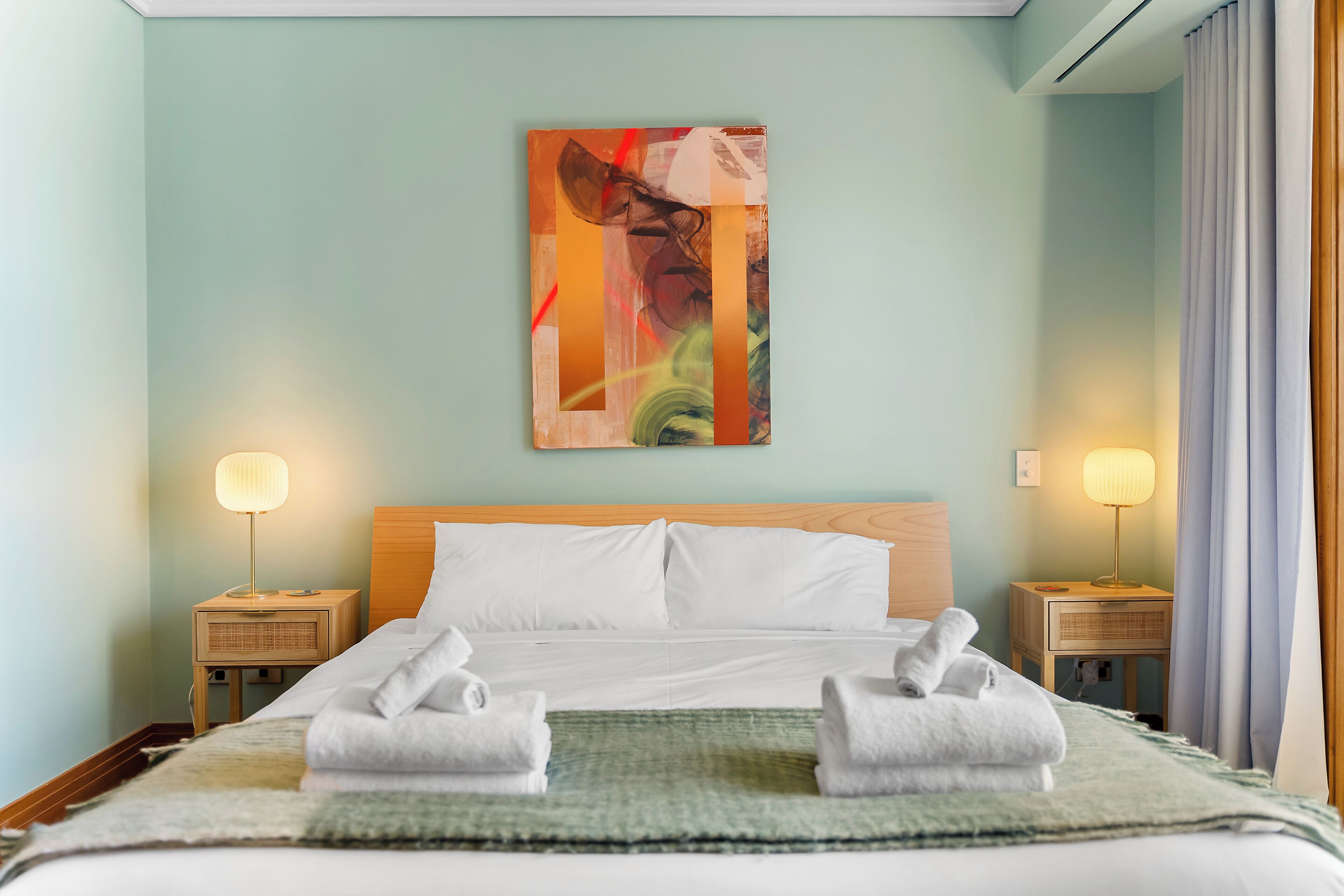 Quarto de hotel com cama, duas mesas de cabeceira, um quadro na parede e uma janela com cortinas.