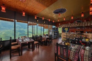 Restaurante com pisos, mesas e cadeiras de madeira, grandes janelas com vista para as montanhas e lanternas penduradas.