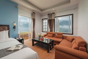 Quarto de hotel com cama, sofá, mesa de centro e vista para montanhas através de grandes janelas.