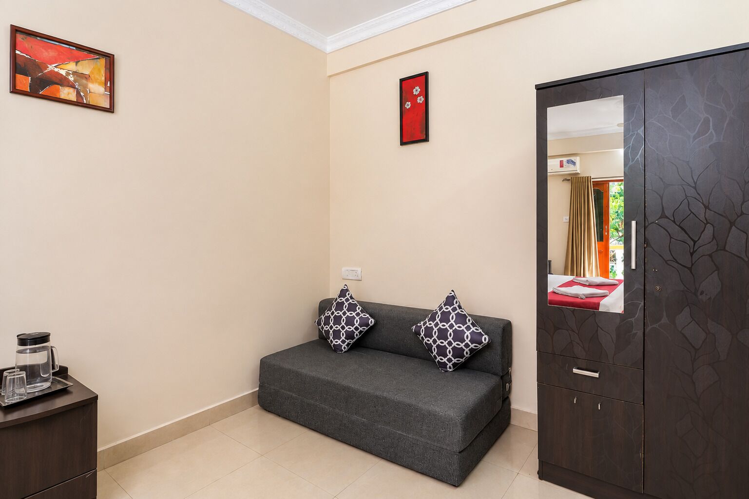Um quarto com um sofá cinzento, duas almofadas estampadas, um guarda-roupa de madeira escura, uma pequena mesa de cabeceira com um recipiente de vidro e um quadro abstrato emoldurado na parede.
