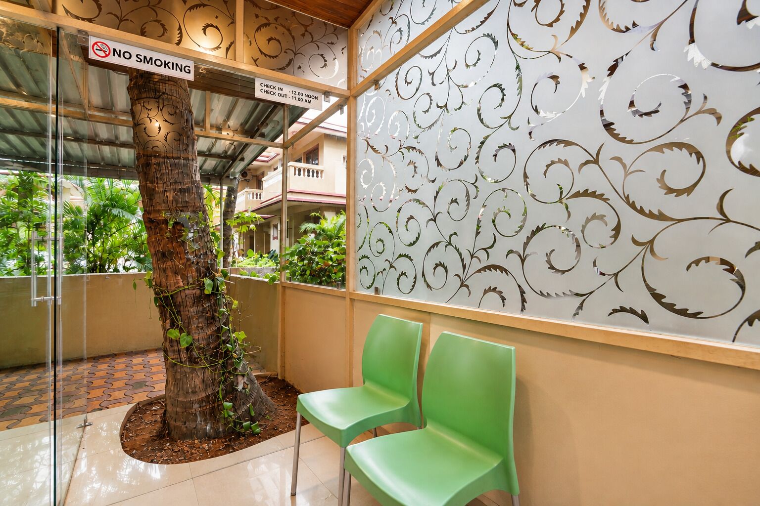 Sinal de proibido fumar, sinal de check-in/check-out, árvore com plantas trepadoras e cadeiras verdes numa área de espera.