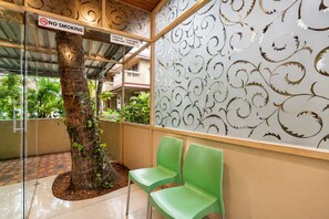 Sinal de proibido fumar, sinal de check-in/check-out, árvore com plantas trepadoras e cadeiras verdes numa área de espera.