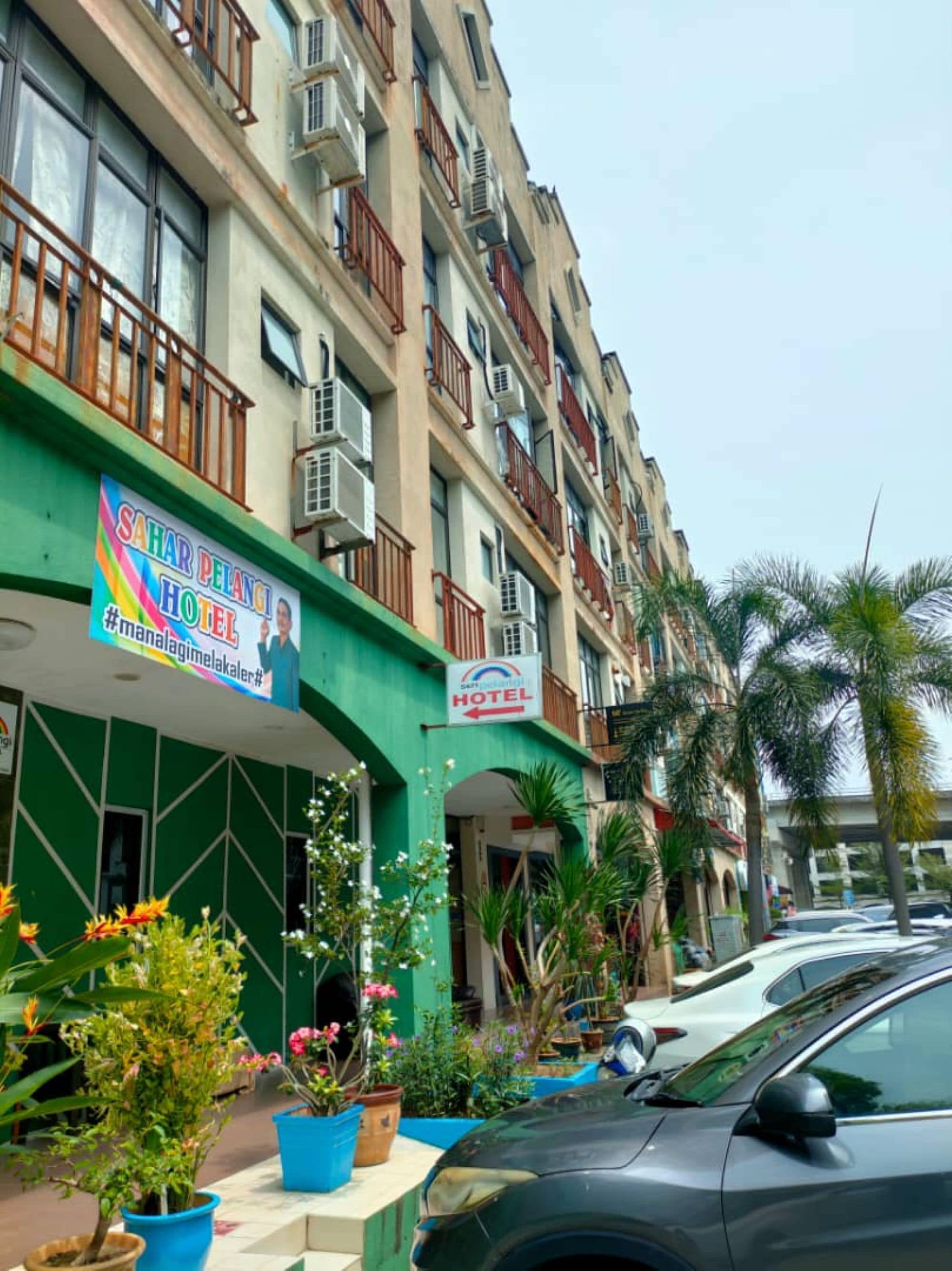 Um hotel com fachada verde, várias varandas e plantas em vasos na área de entrada.