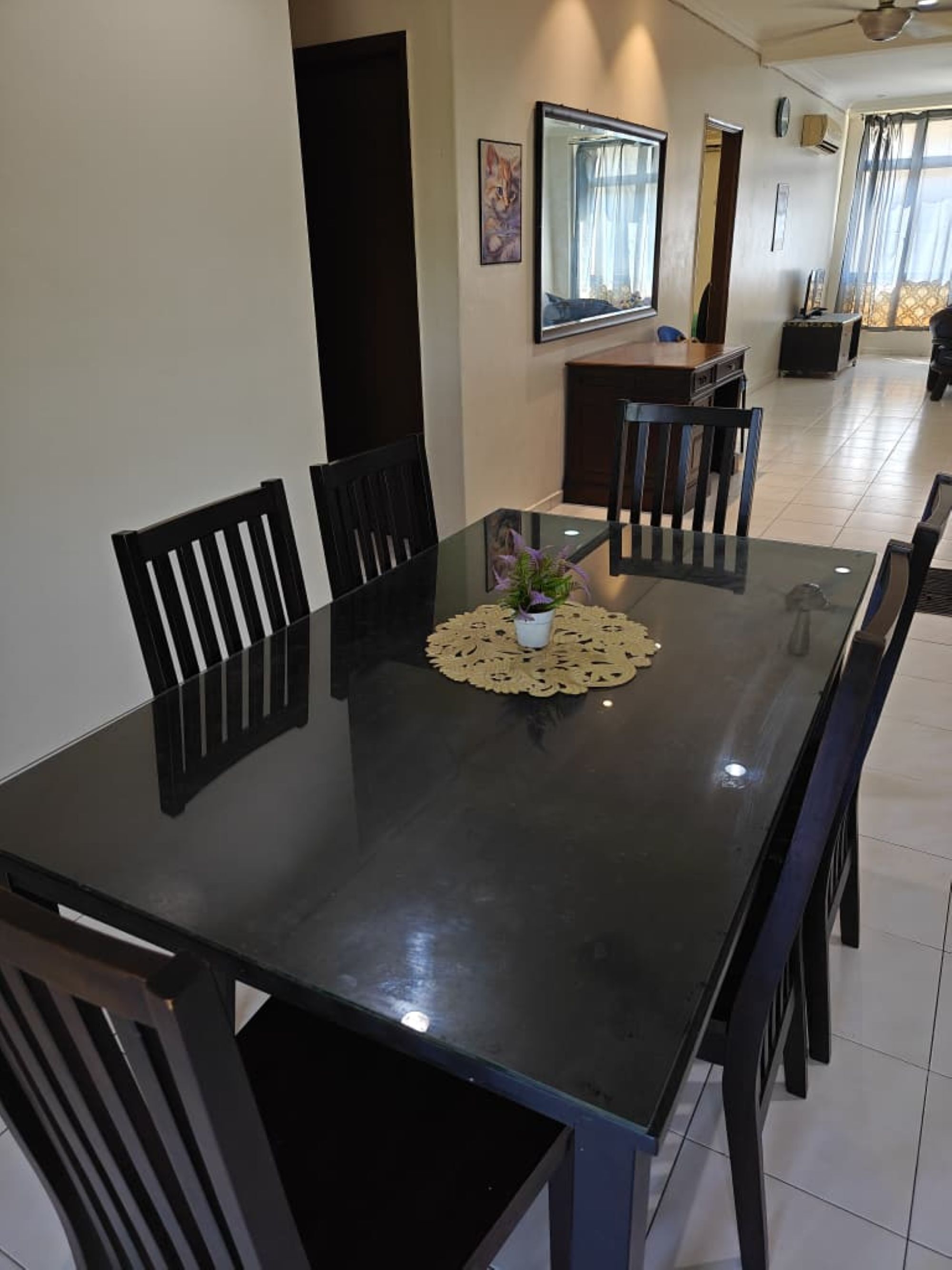 Uma sala de jantar com uma mesa retangular, cadeiras pretas, uma pequena planta em vaso e um jogo americano decorativo.