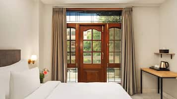 Quarto de hotel com uma cama grande, porta de madeira com painéis de vidro, uma pequena mesa com chaleira e mesa de cabeceira com abajur.