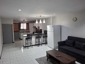 Apartamento moderno com cozinha, sala de jantar e sala de estar. Possui geladeira, máquina de café e sofá.