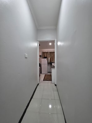 Um corredor estreito com paredes brancas e chão de azulejos leva a uma área de cozinha com geladeira e micro-ondas.