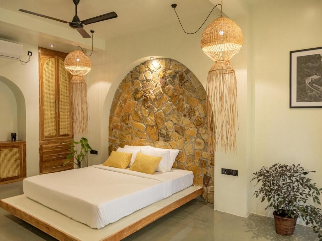 Quarto com parede de pedra, cama com roupa de cama branca, ventilador de teto e duas luminárias pendentes.
