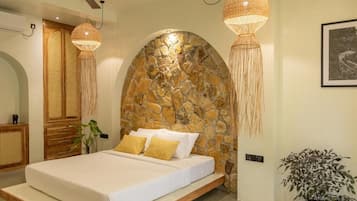 Quarto com parede de pedra, cama com roupa de cama branca, ventilador de teto e duas luminárias pendentes.