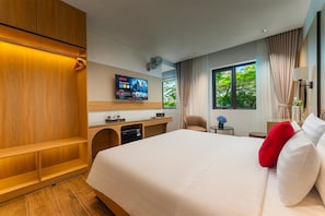 Quarto de hotel com cama grande, escrivaninha, televisão de tela plana, área de estar e janela com vista para árvores.
