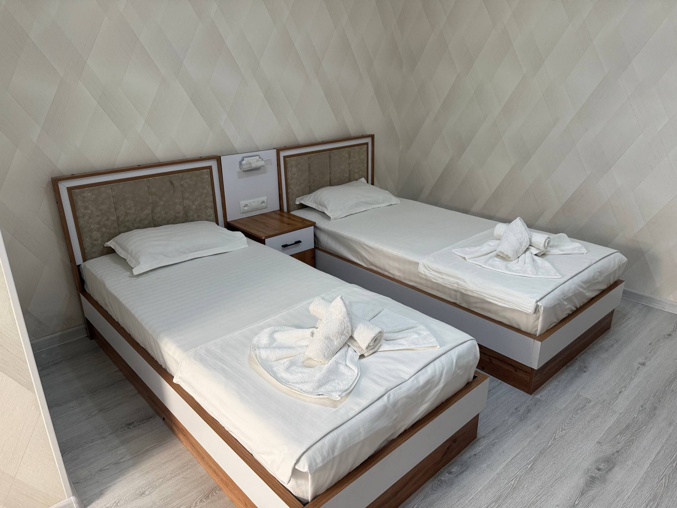 Quarto de hotel com duas camas, cada uma com estrutura de madeira e roupa de cama branca. Há mesas de cabeceira de madeira com gavetas e um aparelho de luz fixado na parede entre as camas.