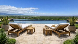 Área à beira da piscina com duas espreguiçadeiras, uma mesa pequena e um vaso de planta.