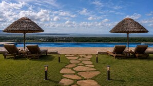 Área da piscina com duas espreguiçadeiras sob toldos de palha, um caminho de pedra e vista para um corpo d'água e paisagem distante.