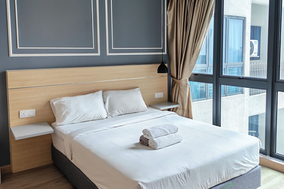 Quarto de hotel moderno com uma cama grande, mesa de cabeceira, cortinas e vista para o exterior.