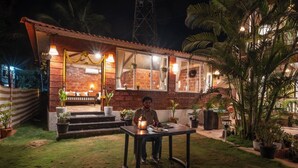 Uma casa de tijolos com varanda, vasos de plantas e uma mesa onde uma pessoa está sentada.