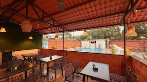 Uma área externa com mesas e cadeiras, piscina ao fundo e uma cobertura.