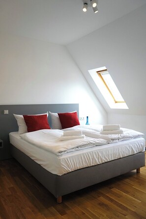 Quarto moderno com teto inclinado, claraboia, cama com roupa de cama branca e cabeceira cinza.