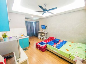 Quarto compacto com cama, escrivaninha, ventoinha no teto e pequena cozinha.