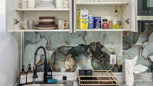 Cozinha com painel de parede em mármore, pia de aço inoxidável e armários abertos com louças e utensílios de cozinha.