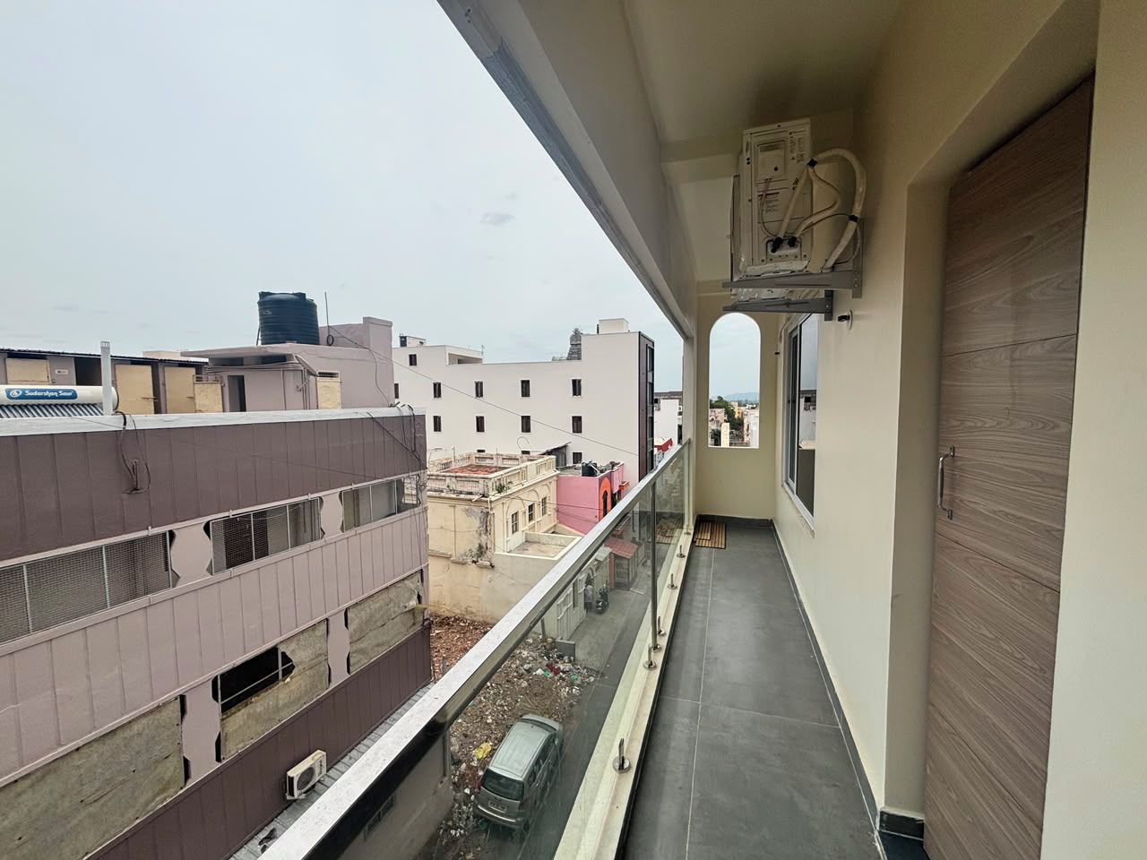 Uma varanda com vista para edifícios próximos, uma janela com persianas e uma porta com painel de madeira.