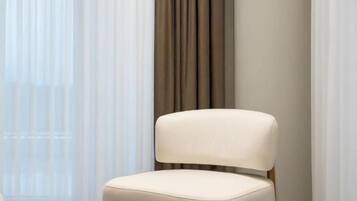 Quarto com uma única cadeira, janela com cortinas translúcidas e um painel vertical na parede.