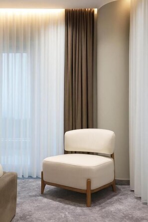 Quarto com uma única cadeira, janela com cortinas translúcidas e um painel vertical na parede.