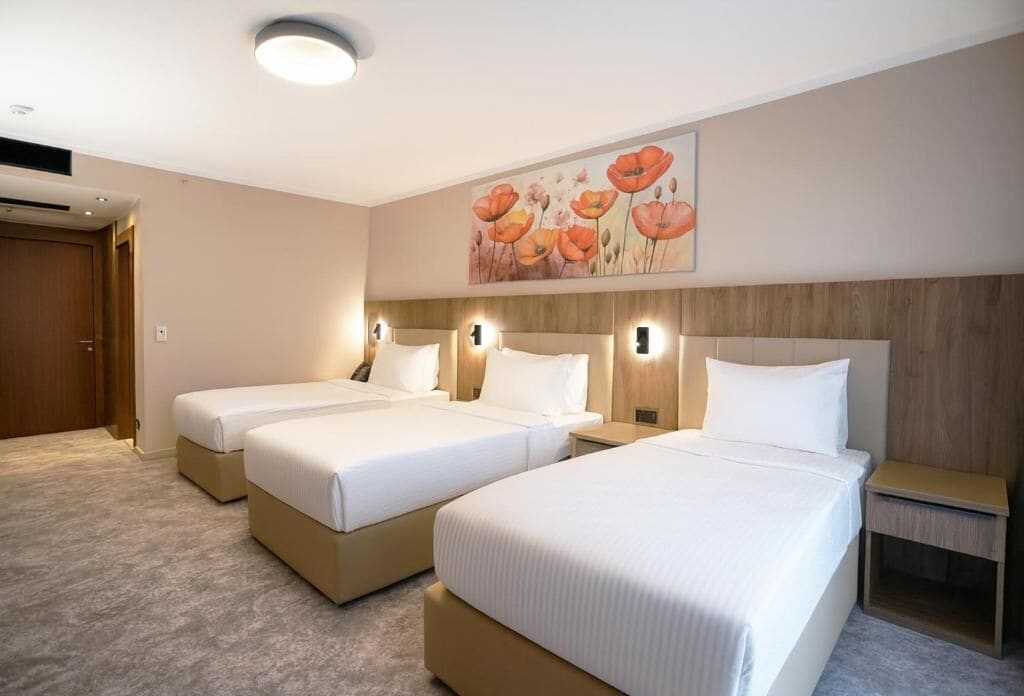 Quarto de hotel com três camas, cabeceira de madeira, pintura de flores e piso coberto por carpete.