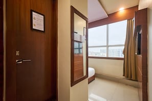 Quarto de hotel com porta de madeira, um espelho grande, janela com vista para edifícios e uma cama com lençol branco.
