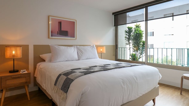 Quarto de hotel com uma cama grande, duas luminárias de cabeceira, um quadro na parede e uma janela com vista para o exterior.