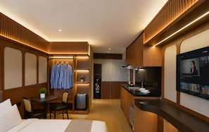 Quarto de hotel com cama, escrivaninha, cadeira, pequena mesa com planta, mala, cozinha compacta e televisão exibindo um vídeo.