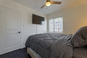 Quarto com cama, ventilador de teto, televisão fixada na parede e janela com persianas.