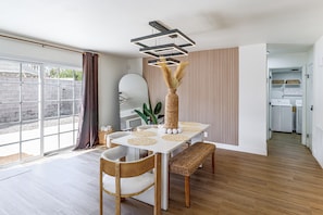 Uma sala de jantar com mesa branca, cadeiras de fibra natural, um vaso decorativo e uma luminária pendente moderna.