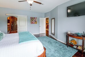 Quarto com cama, ventoinha no teto, televisão e porta que dá para um banheiro.