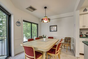 Uma área de refeições com uma mesa grande, cadeiras, luminária pendente e janela com vista para árvores.