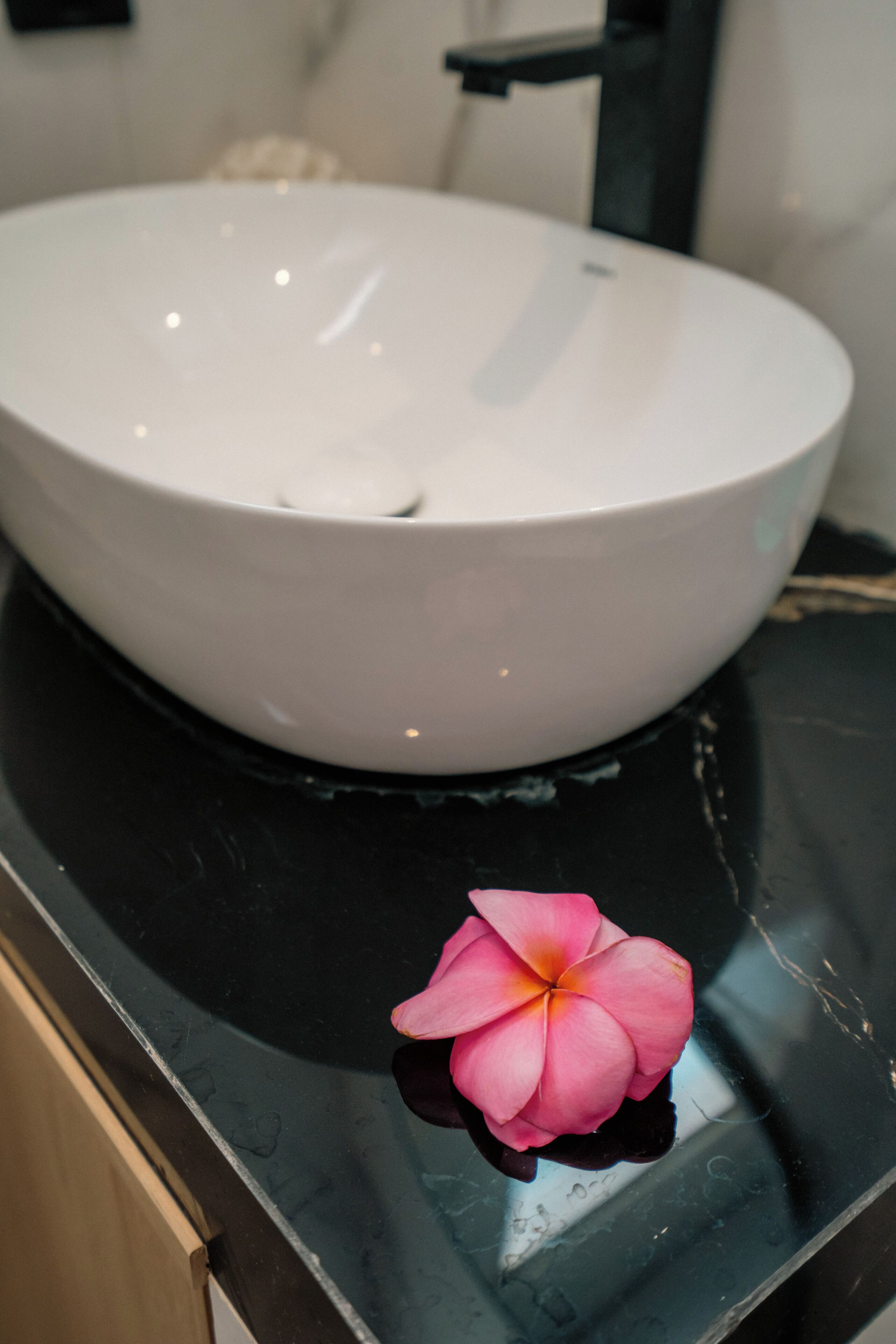 Uma pia branca sobre bancada preta, com uma única flor rosa ao lado.