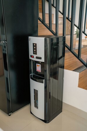 Uma geladeira compacta e moderna com painel de controle digital, ao lado de um pilar preto em um quarto com piso de madeira e uma escada ao fundo.