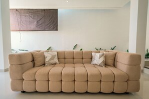Um sofá modular bege com almofada decorativa estampada, posicionado em um quarto com parede branca lisa e janela com persiana marrom.