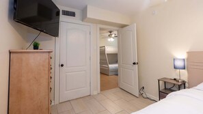 Quarto com cama, televisão na parede, mesa de cabeceira com luminária e porta que dá para um banheiro com beliche.