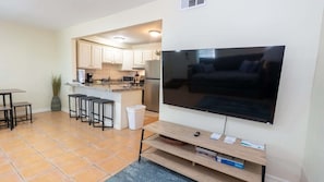 Cozinha moderna com armários brancos, uma grande TV de tela plana na parede, área de jantar com mesa e banquetas, e uma pequena lixeira.