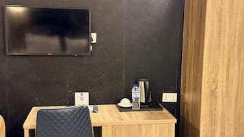 Quarto de hotel com televisão de tela plana fixada na parede, escrivaninha de madeira com cadeira, garrafa de água, chaleira e mesa pequena com prato e xícara.