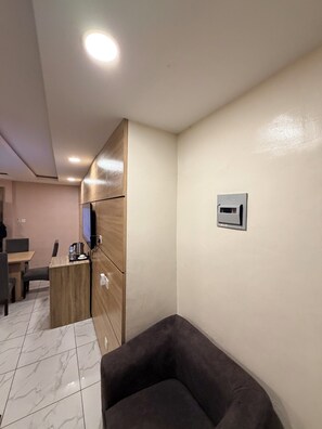 Quarto de hotel com área de estar, uma mesinha com chaleira, televisão e dispositivo fixado na parede.